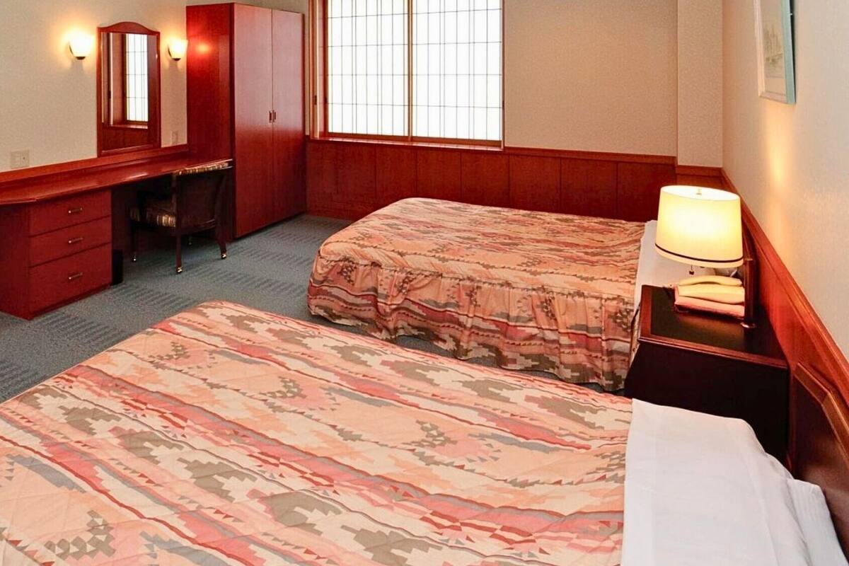 Quarto de hotel com duas camas, uma escrivaninha, uma cadeira, um guarda-roupa e uma janela com persianas.