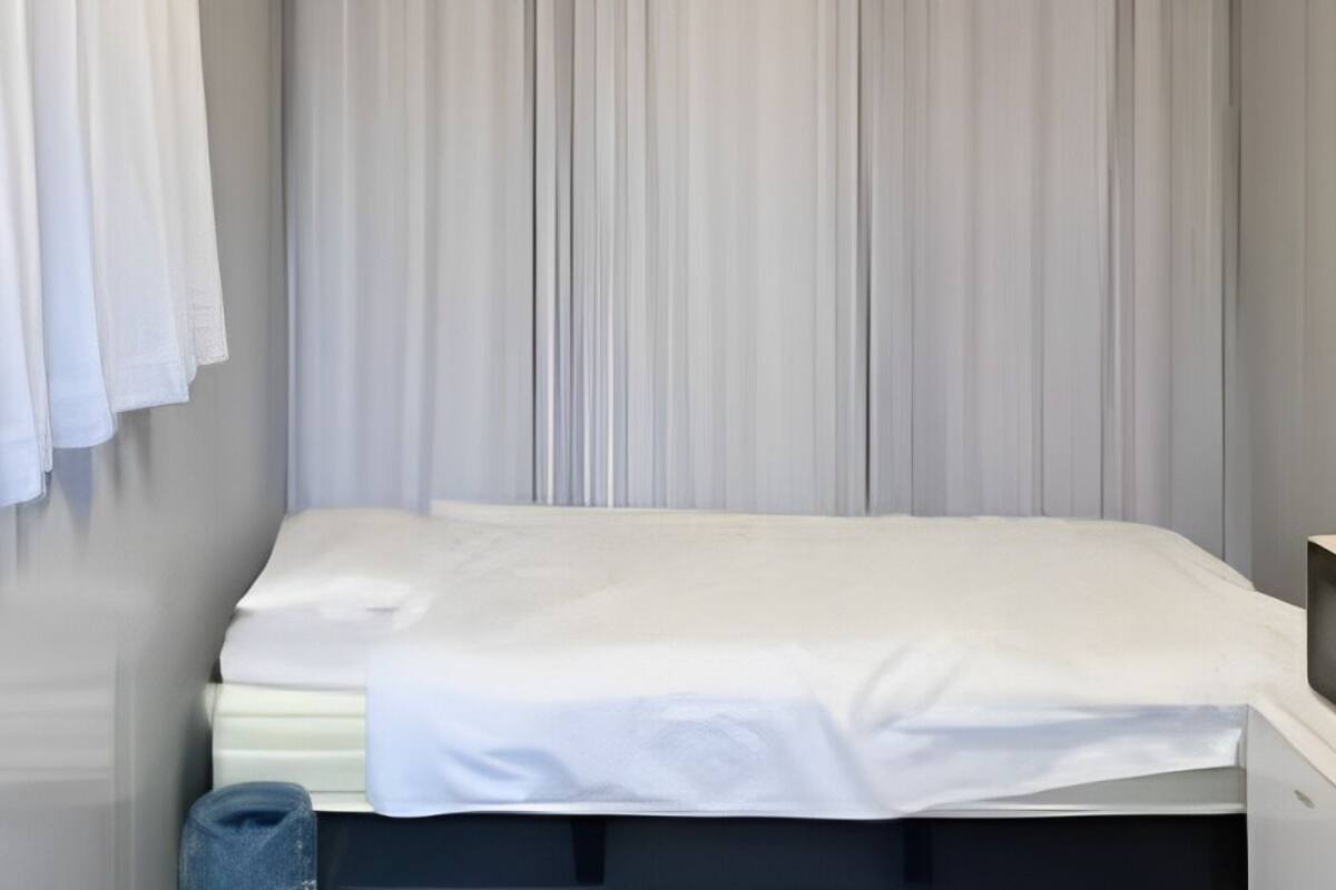 Uma cama de solteiro com colchão e lençóis brancos, uma mesinha de cabeceira, uma televisão fixada na parede e uma janela com cortinas brancas.