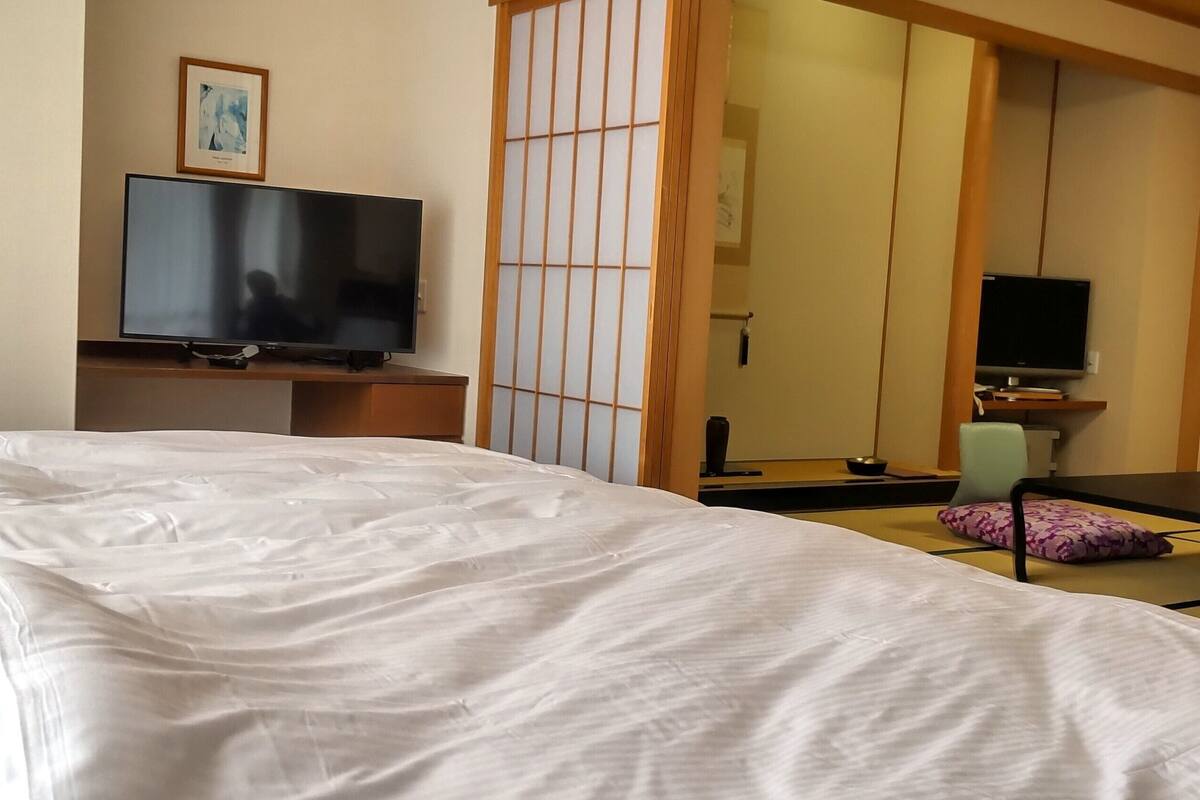 Quarto de hotel com cama, televisão em suporte, uma cadeira e um espelho.