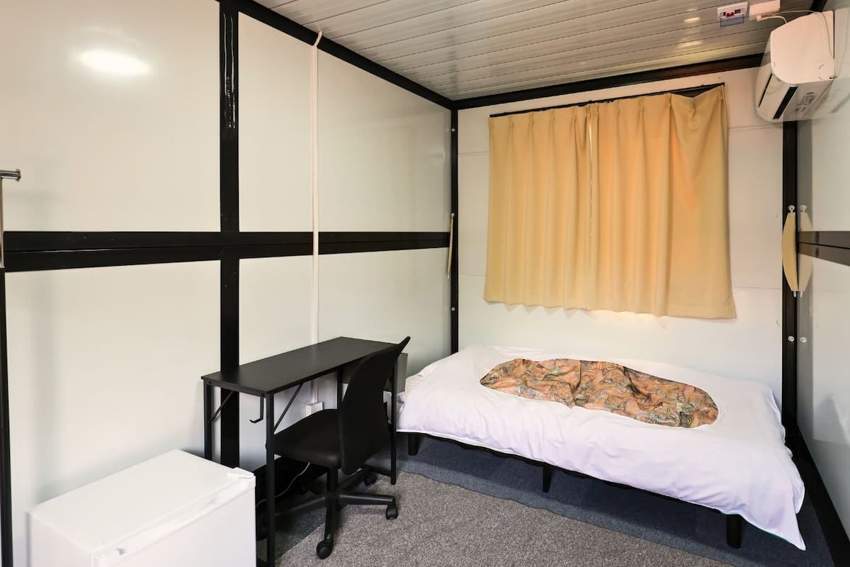 Quarto pequeno e compacto com uma cama de solteiro, uma escrivaninha e uma cadeira.