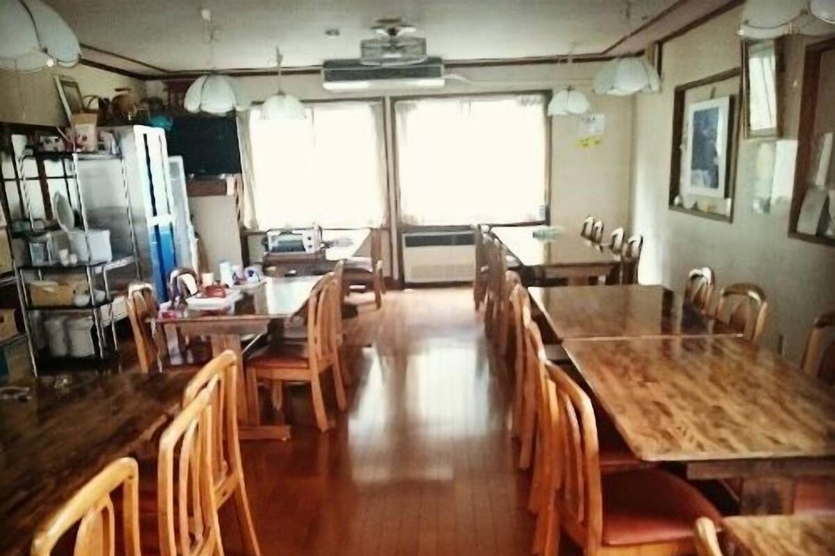 Uma área de refeições com mesas e cadeiras de madeira, uma cozinha ao fundo e grandes janelas.