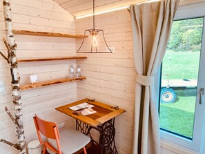 Quarto aconchegante com paredes de madeira, uma mesa com uma revista, uma cadeira, uma luminária pendente e uma janela com vista para um trator.