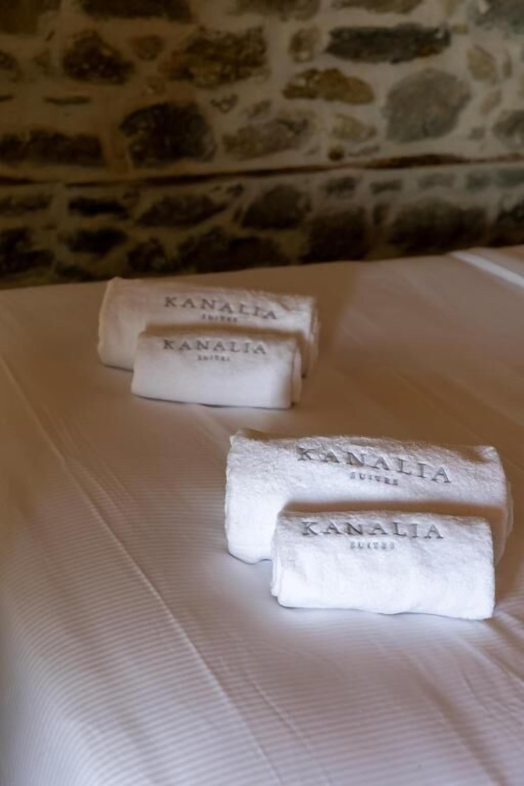 Cama com toalhas dobradas com cuidado, identificadas com 'KANALIA', e parede de pedra ao fundo.