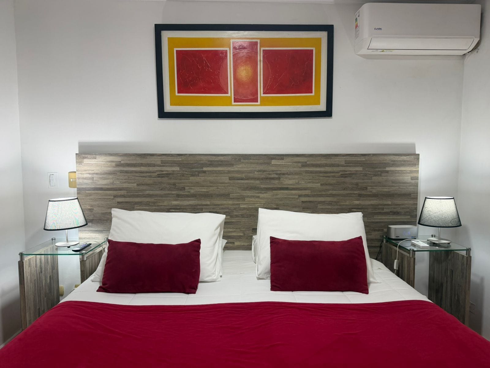 Quarto de hotel com cabeceira de madeira, duas mesas de cabeceira com luminárias, cama com roupa de cama branca e vermelha e uma obra de arte emoldurada acima da cama.