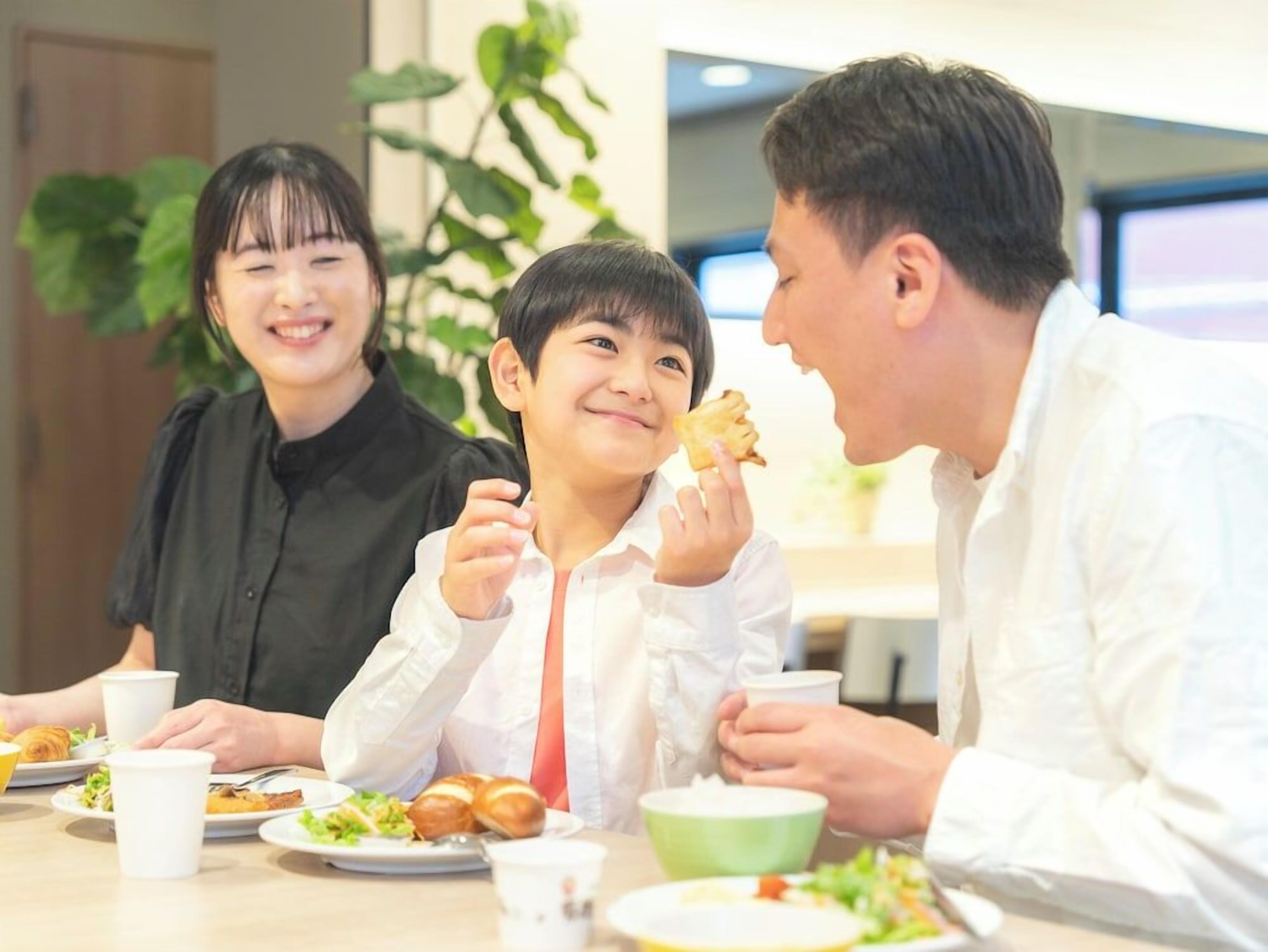 Um grupo de pessoas sentadas à mesa, com comida e bebidas, num espaço com uma planta e uma janela.