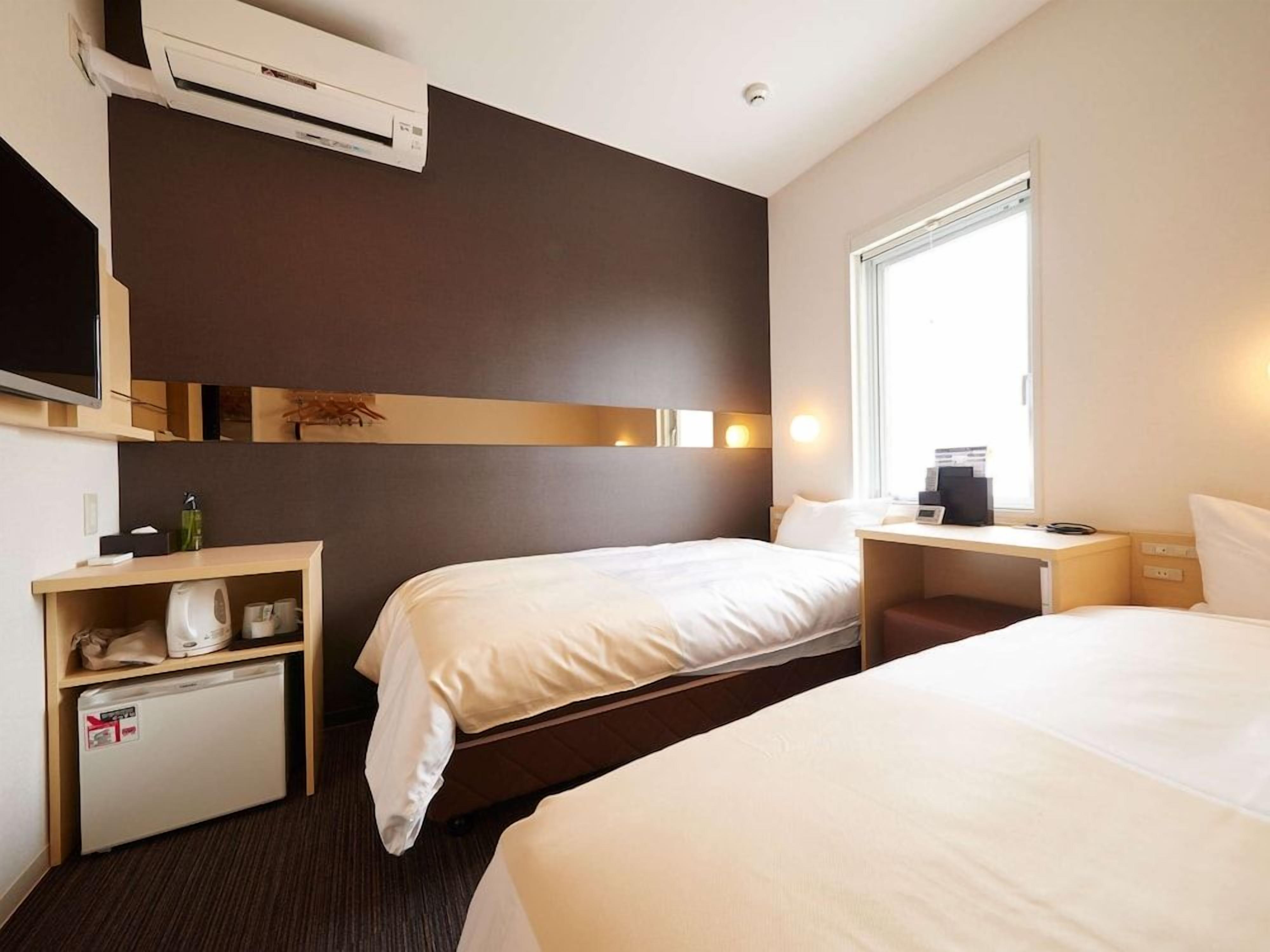 Quarto de hotel com duas camas, uma escrivaninha, um mini-refrigerador, uma televisão e uma janela.