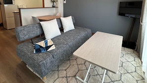 Uma sala de estar moderna com um sofá cinzento, uma mesa de centro de madeira, uma cozinha com micro-ondas e uma TV fixada na parede.