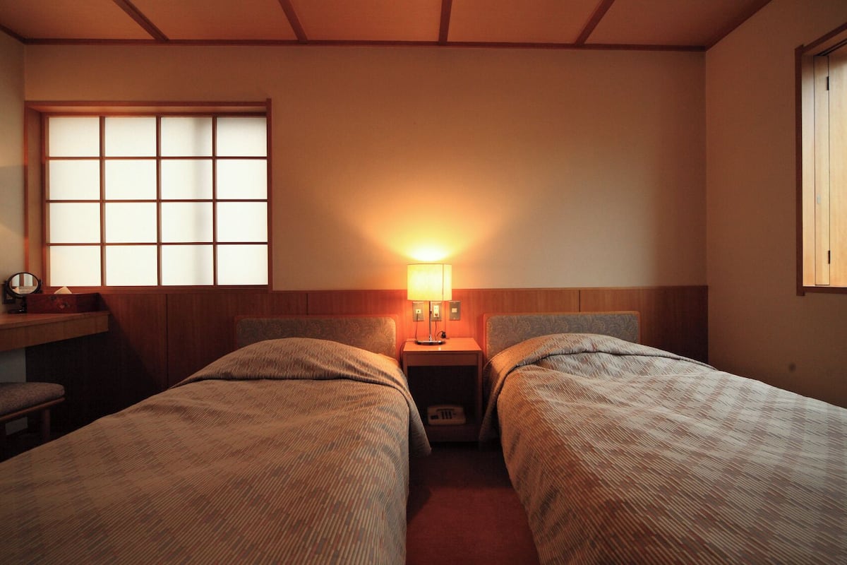 Quarto com duas camas, uma mesa de cabeceira com abajur, janela com vista e escrivaninha com espelho.