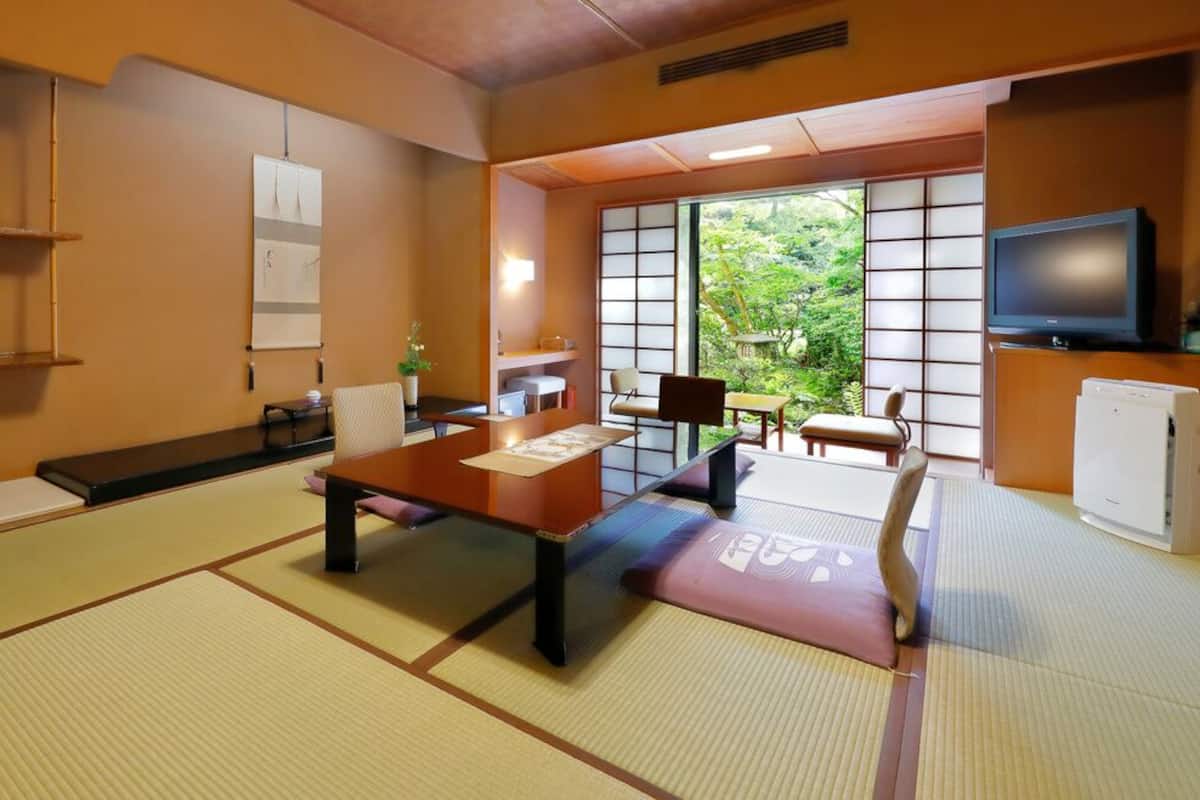 Um quarto japonês tradicional com tatames, uma mesa baixa, cadeiras, televisão e vista para o verde lá fora.