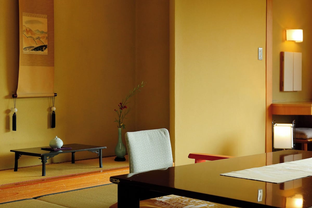 Quarto de hotel com uma mesa de madeira, uma cadeira, uma pequena escrivaninha, um vaso com flores e uma prateleira com luminária.