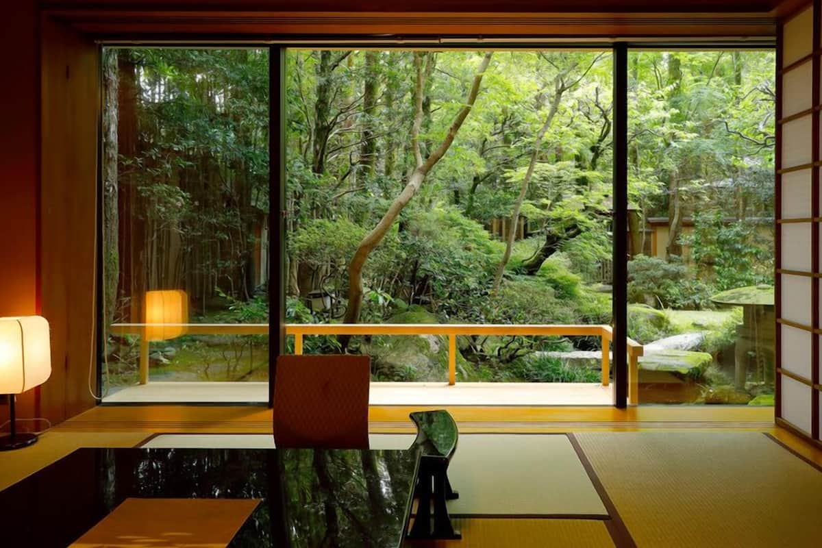 Um quarto japonês tradicional com tatames, uma mesa baixa, um banco e uma janela grande com vista para um jardim arborizado.