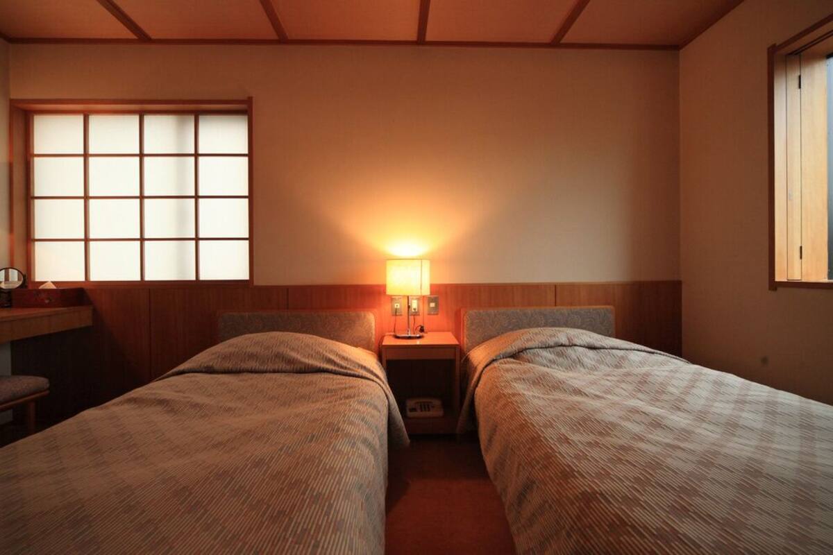 Quarto com duas camas, janela com vista, mesa de cabeceira com abajur e um pequeno banco.