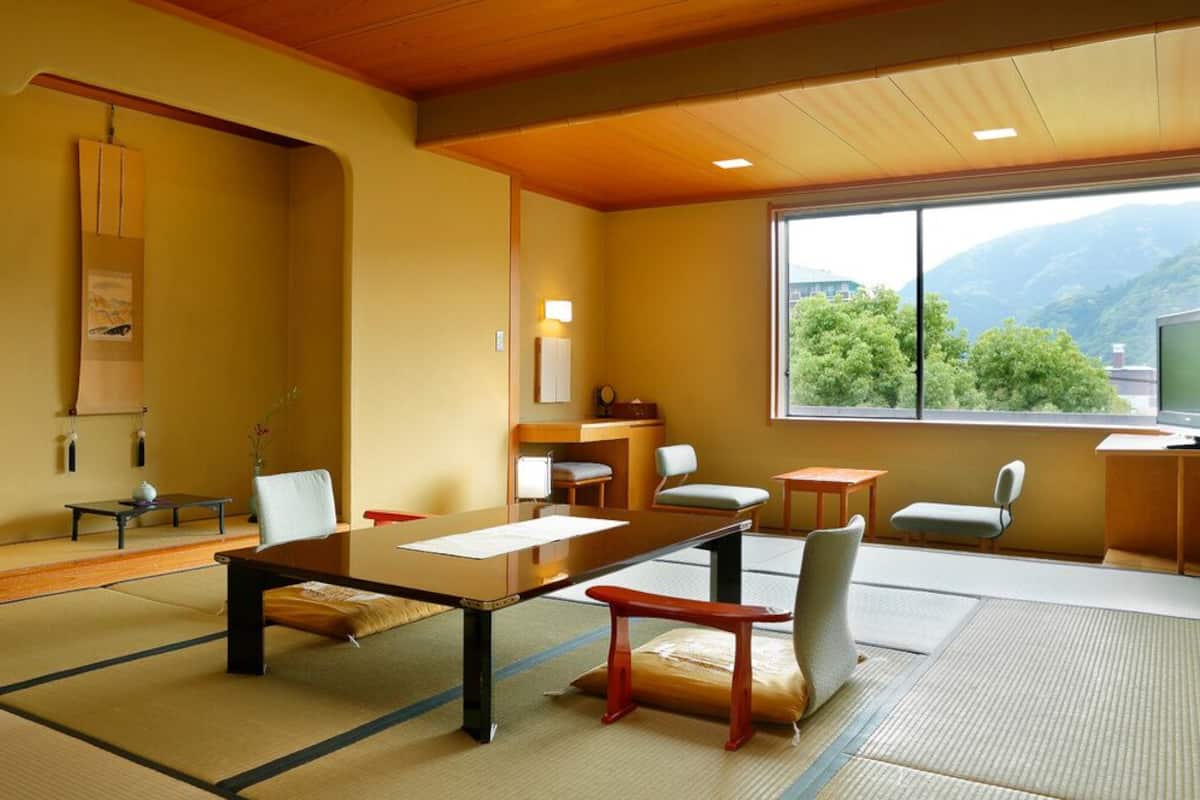 Um quarto japonês tradicional com tatames, uma mesa baixa, cadeiras, uma televisão e uma janela com vista para o verde.