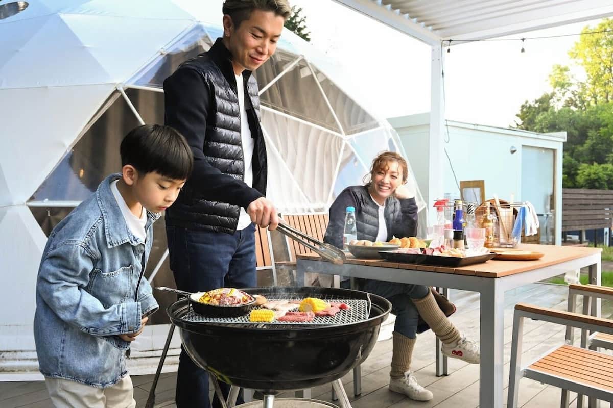 Uma família fazendo churrasco ao ar livre, com uma estrutura de cúpula geodésica transparente ao fundo.