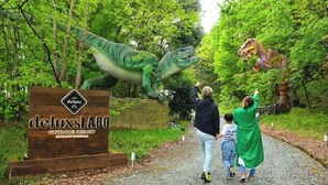 Um letreiro de madeira com os textos 'deluxsLABO' e 'OUTDOOR RESORT', rodeado por vegetação e estátuas de dinossauros.