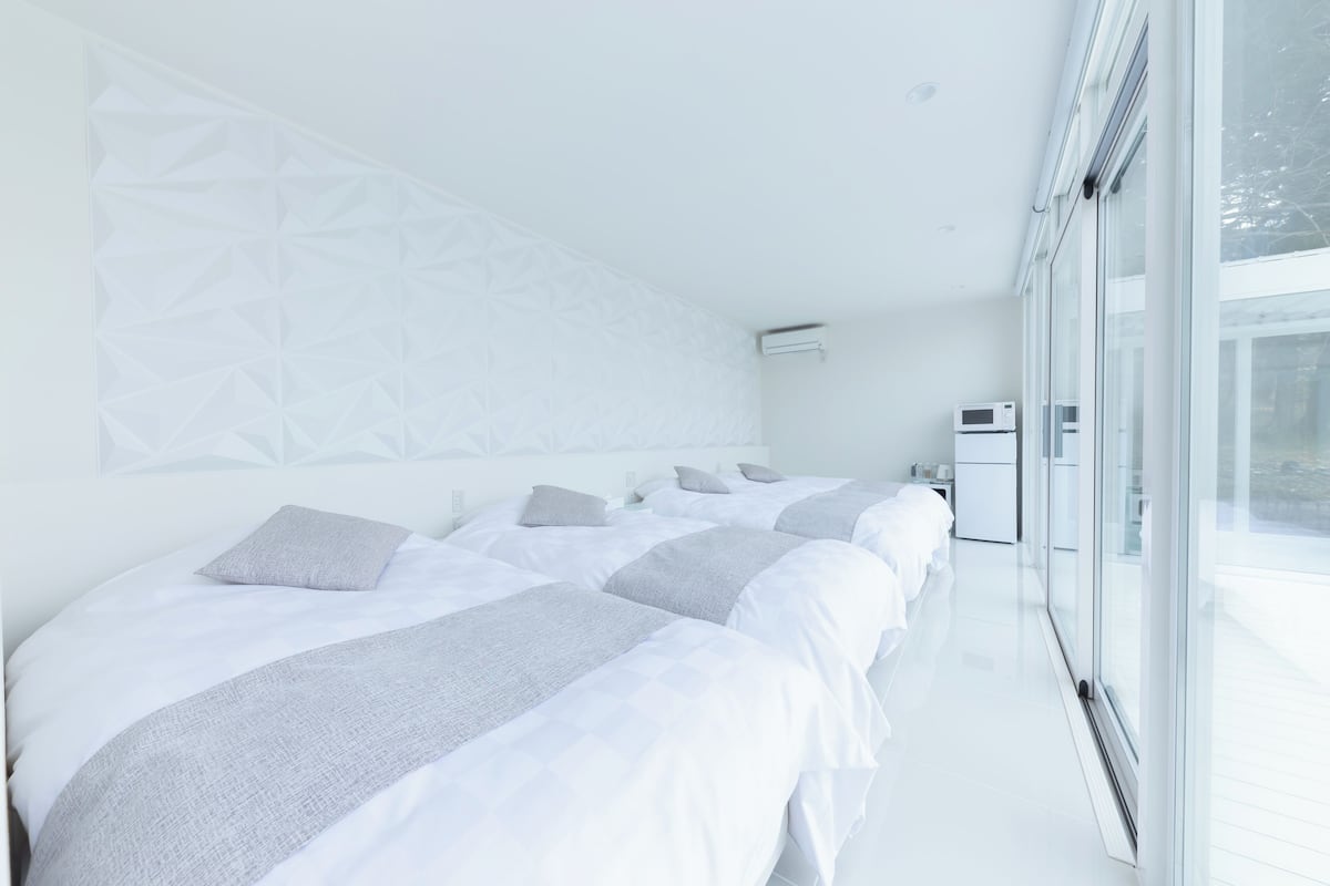 Quarto de hotel moderno com design geométrico na parede, uma janela grande e uma cama com manta cinza.