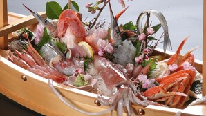 Barco de madeira repleto de mariscos variados, incluindo camarão, peixe e lula, dispostos sobre uma base de folhas verdes e decorado com flores.