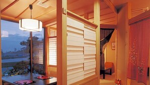 Um quarto japonês tradicional com piso de tatame, uma porta deslizante, uma mesa com abajur e vista pela janela.