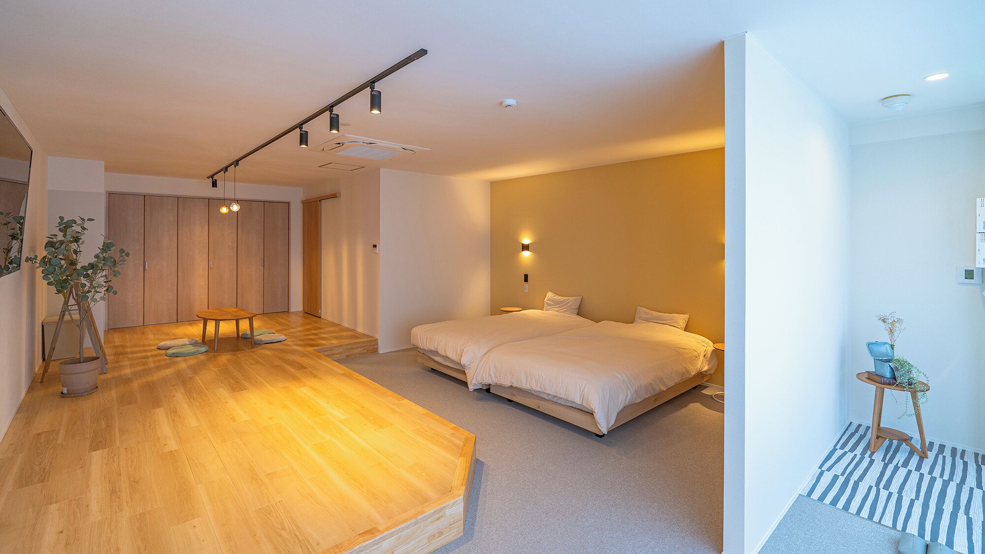 Quarto de hotel moderno com piso de madeira, uma cama grande, uma escrivaninha e uma mesinha com uma planta.