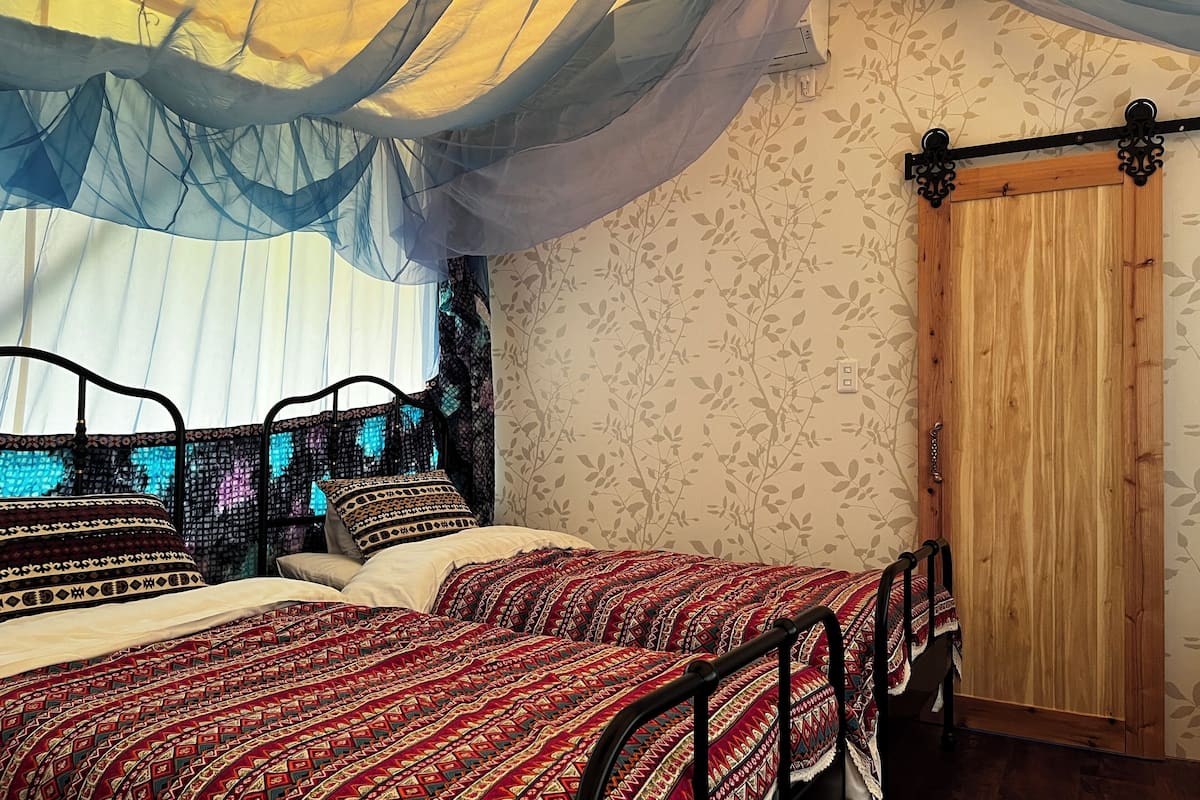Quarto com cama, porta de madeira e janela com cortinas leves.