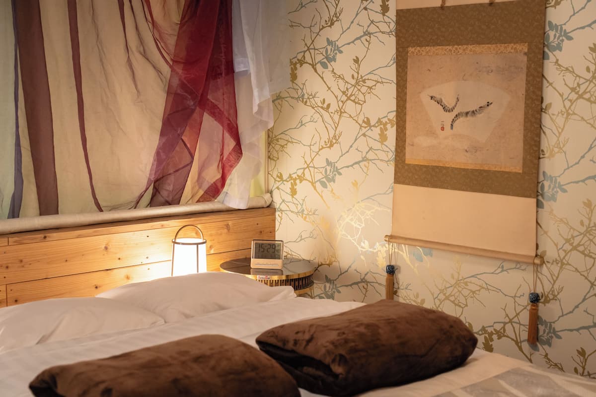 Quarto com cama, almofadas, mesa de cabeceira com relógio, quadro na parede e janela com cortinas leves.
