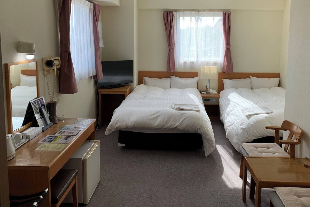 Quarto de hotel com duas camas, uma escrivaninha com computador, uma televisão e uma pequena mesa com cadeira.