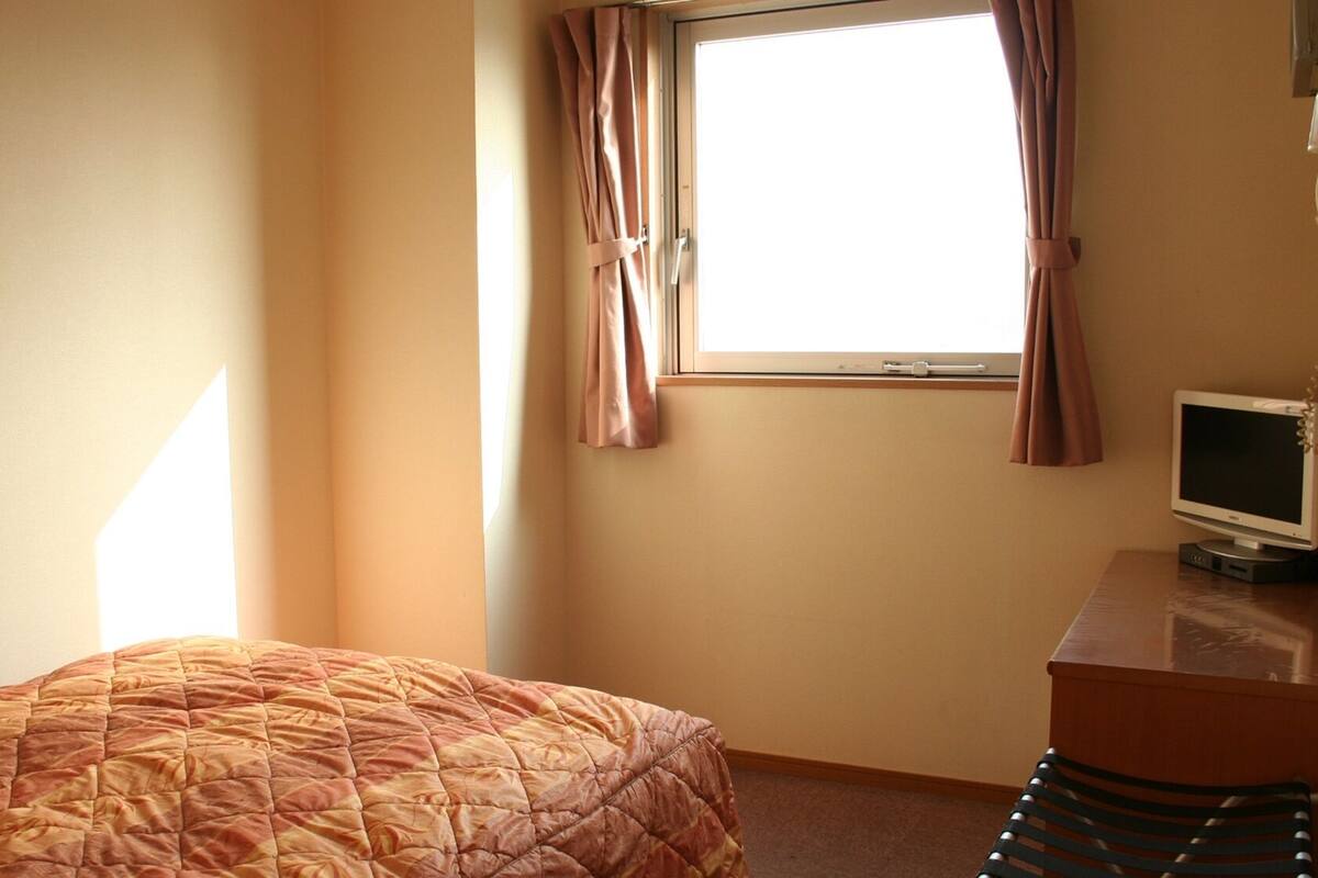 Um quarto com cama, janela com cortinas, televisão sobre suporte e cómoda de madeira.