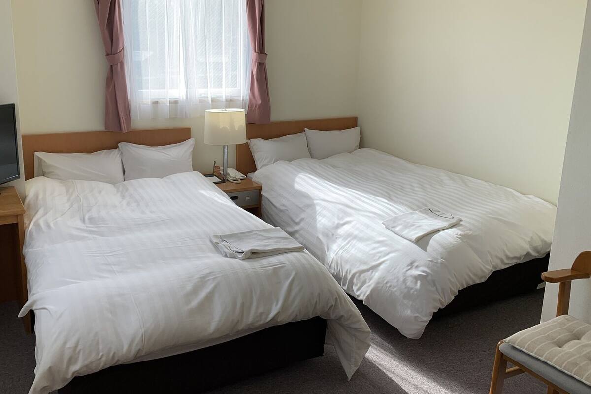 Quarto de hotel com duas camas de solteiro, uma televisão sobre uma mesa de cabeceira de madeira, uma luminária e uma cadeira.