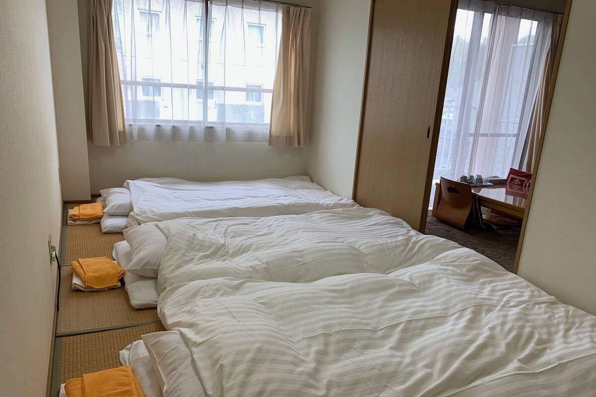 Quarto de hotel com três camas, uma janela com cortinas, uma mesa pequena com uma cadeira e uma porta.
