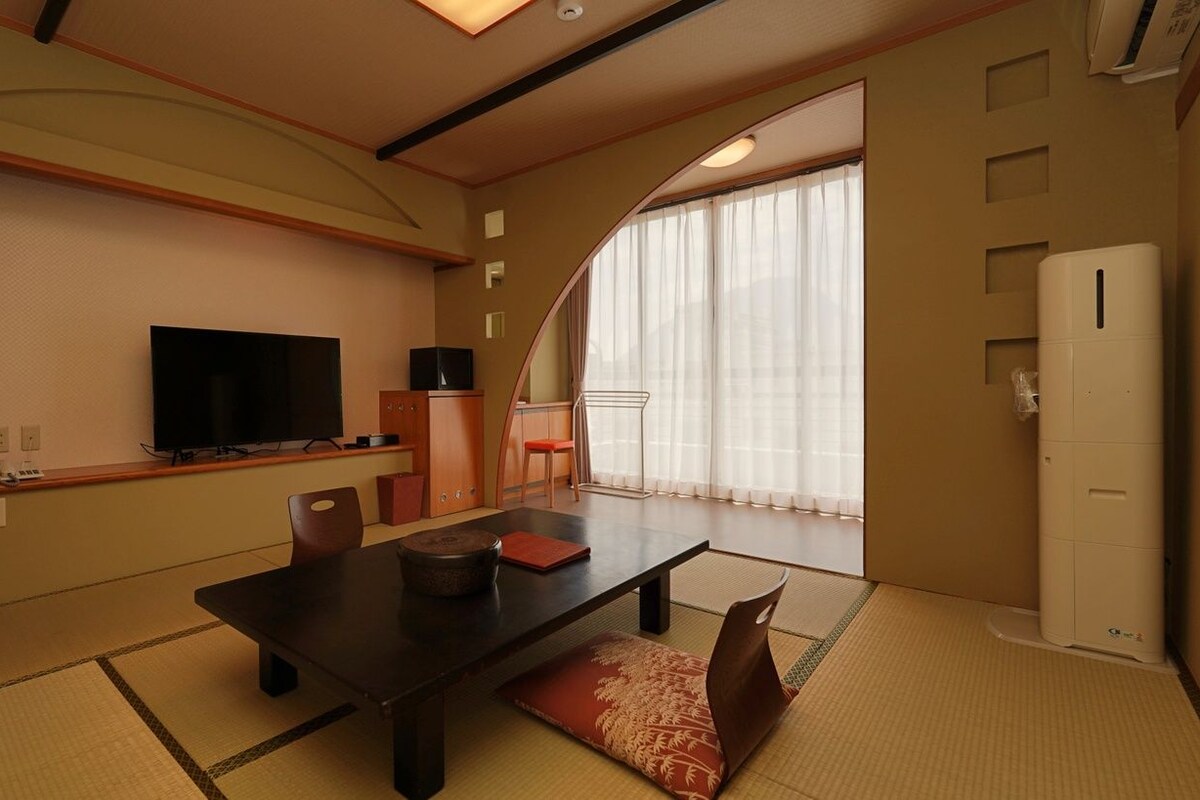 Quarto de hotel moderno com televisão de tela plana, mesa de jantar, sofá e uma grande janela com cortinas.