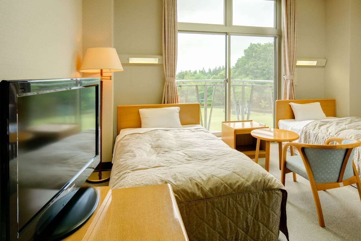 Quarto de hotel com duas camas, televisão, escrivaninha, cadeira e janela com vista para o verde.
