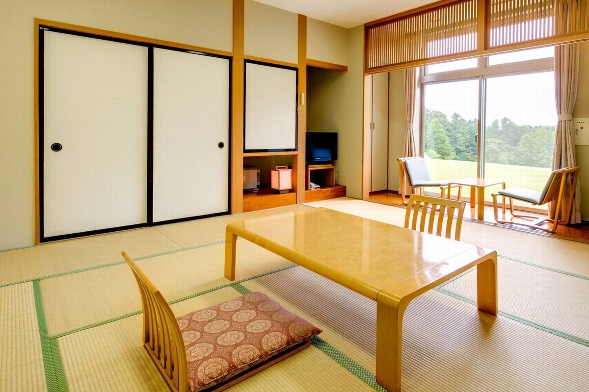 Um quarto japonês tradicional com tatames, uma mesa baixa, cadeiras, televisão e portas deslizantes.
