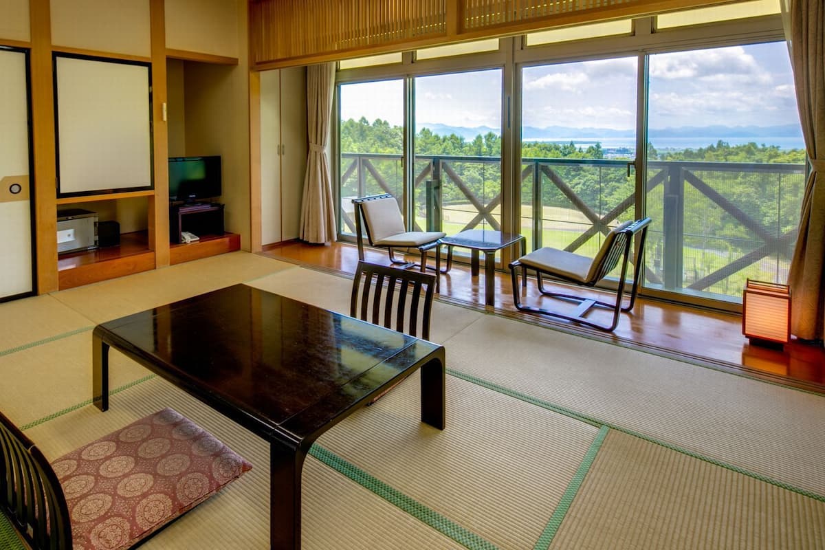 Um quarto tradicional com esteiras de tatame, uma mesa baixa e cadeiras, uma televisão e uma varanda com vista.