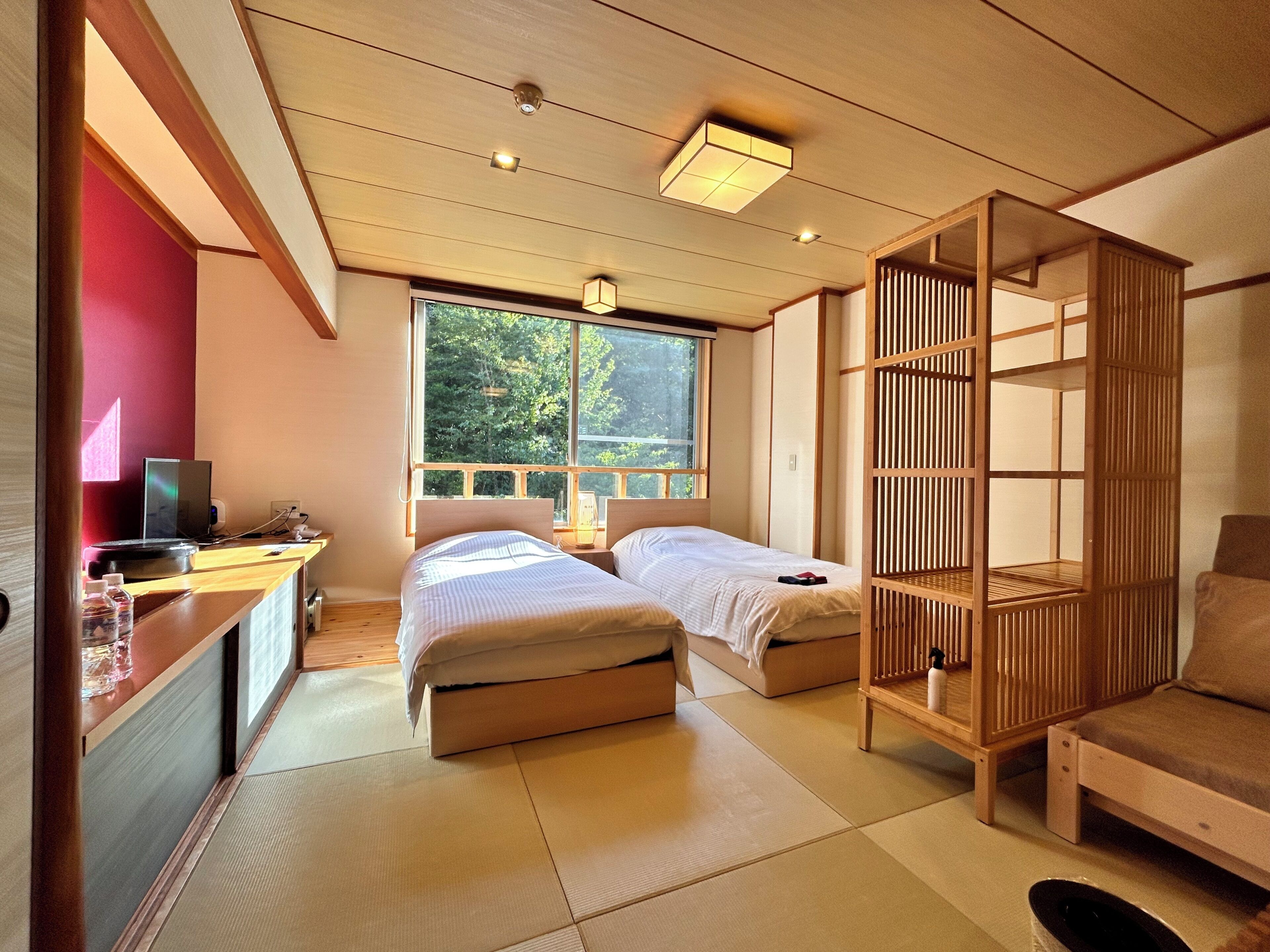 Quarto em estilo japonês tradicional, com tatames, duas camas, uma escrivaninha com computador e um biombos de madeira.