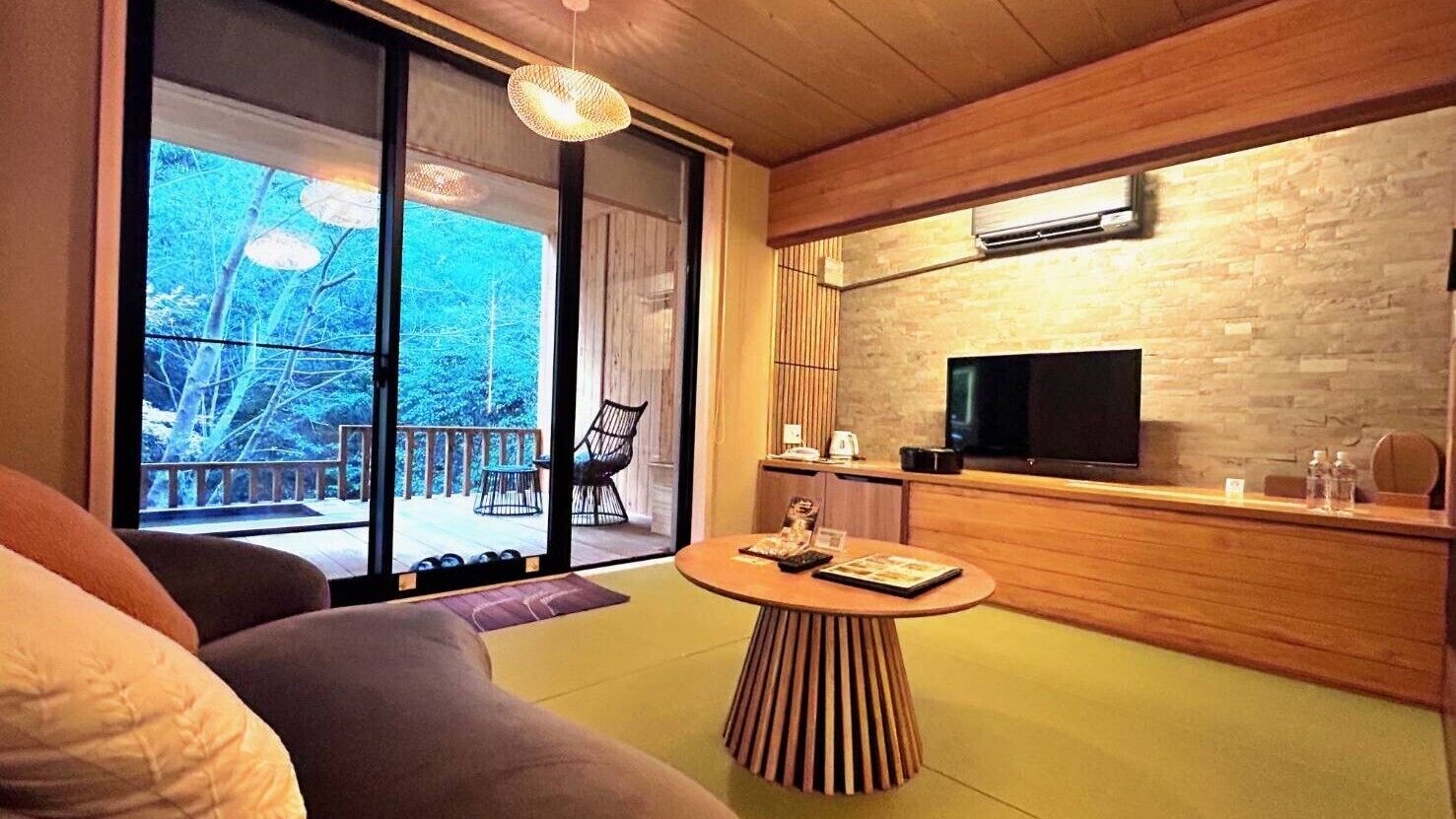 Uma sala de estar moderna com uma grande porta de vidro deslizante que dá para um deck externo, uma mesa de centro redonda de madeira, uma TV de tela plana fixada em uma parede de tijolos e um sofá.
