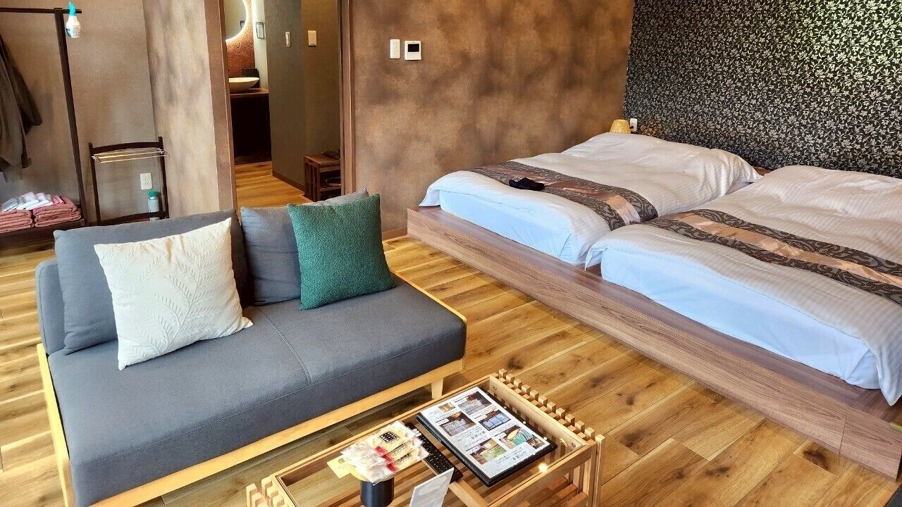 Quarto de hotel moderno com sofá, duas camas, uma mesa de centro de madeira e uma pequena mesa lateral.