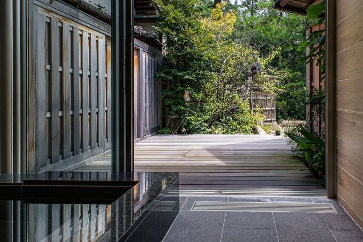 Um edifício tradicional em estilo japonês, com fachada de madeira, um caminho de azulejos que leva a uma porta de madeira e um jardim com árvores e arbustos.