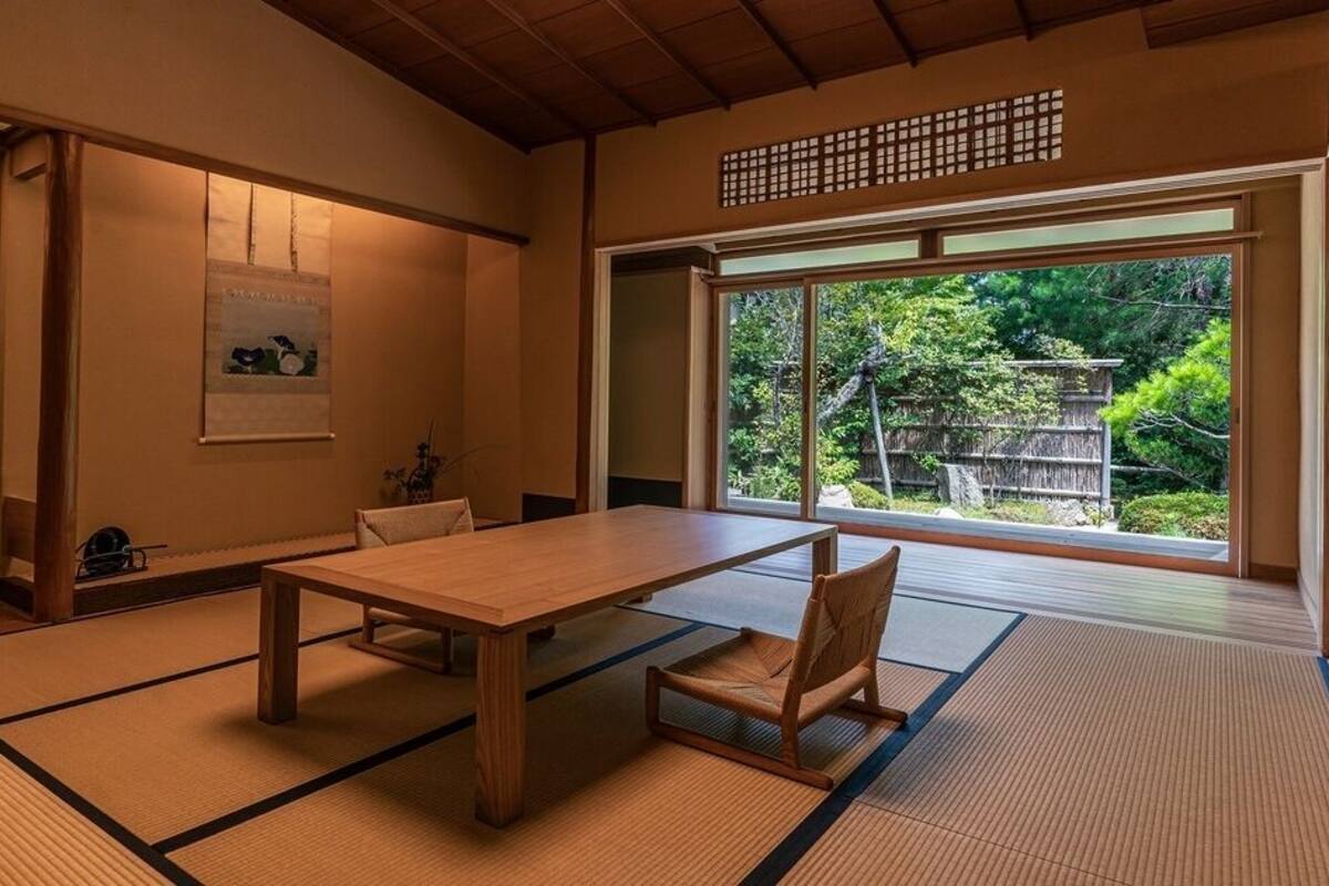 Um quarto japonês tradicional com piso de tatame, uma mesa de madeira, uma cadeira e uma janela grande com vista para o verde.