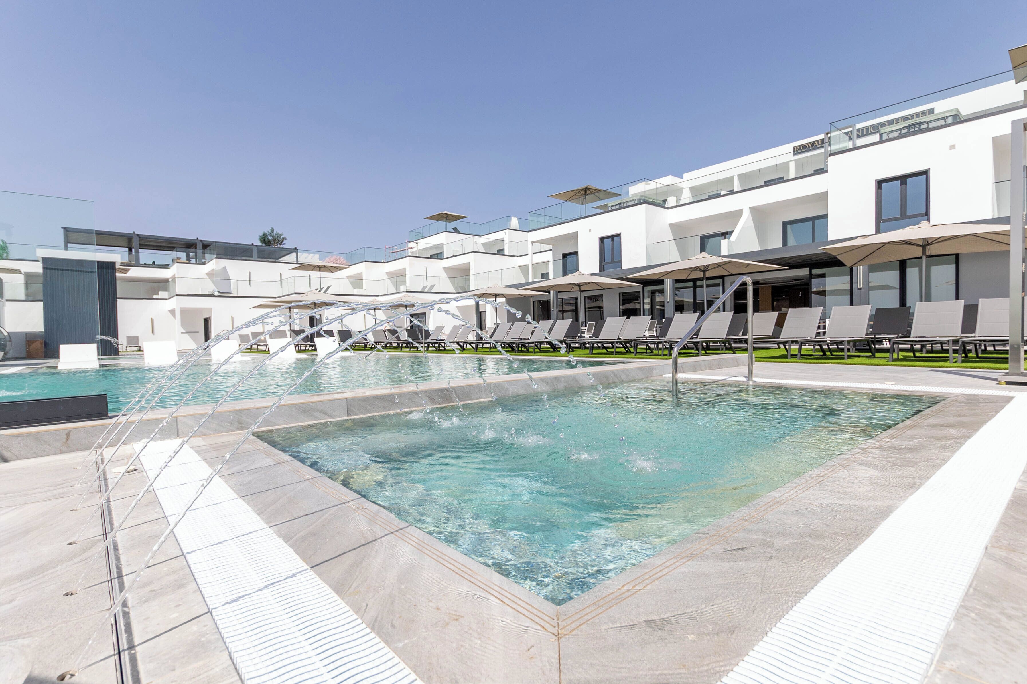 Uma área de piscina com espreguiçadeiras e guarda-sóis, um edifício moderno com varandas e um céu claro.