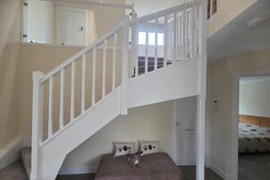 Escadaria com corrimãos brancos que leva a um segundo andar, um quarto com cama e duas almofadas, e uma porta de banheiro.