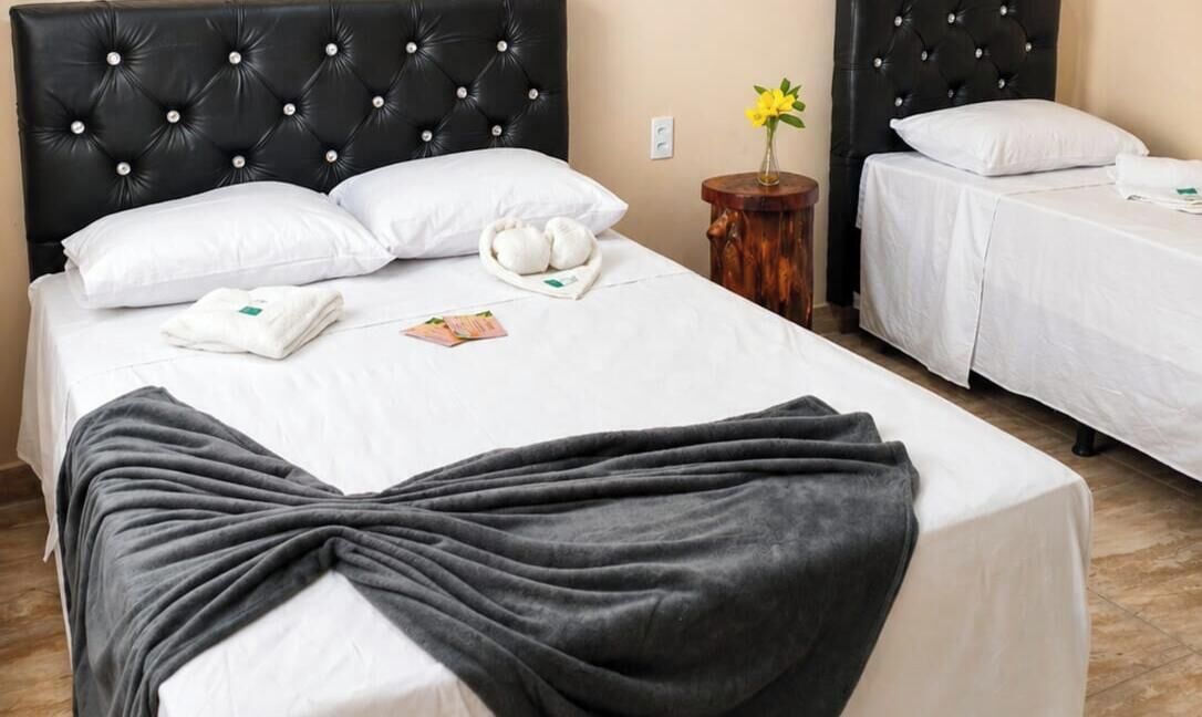 Quarto de hotel com duas camas, cada uma com cabeceira preta estofada, roupa de cama branca e uma pequena mesa de cabeceira de madeira com um vaso de flores amarelas.