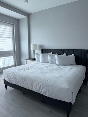 Quarto moderno com uma cama grande, mesa de cabeceira com abajur e janela com persianas.