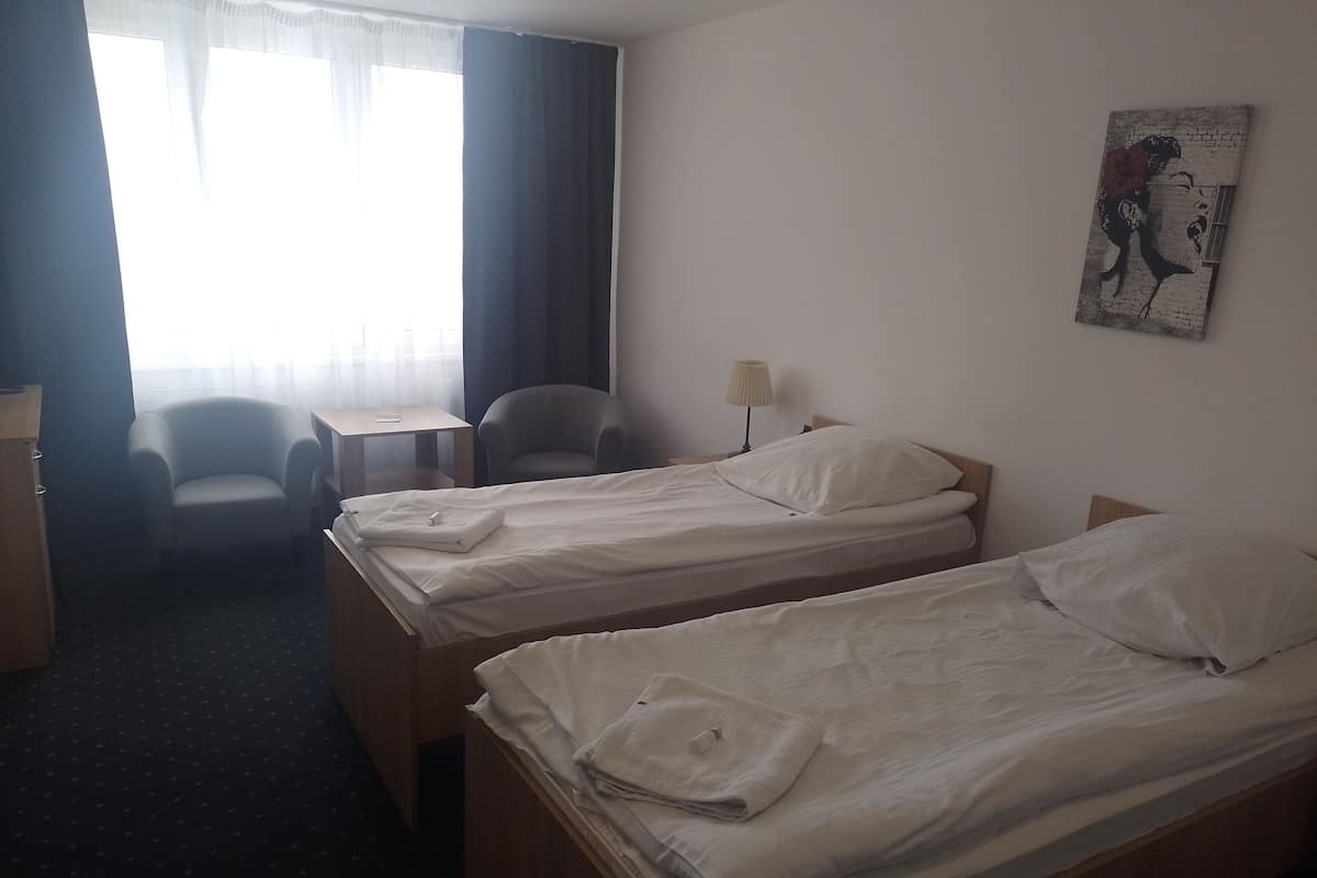 Quarto de hotel com duas camas de solteiro, uma cadeira, uma mesinha e um abajur.