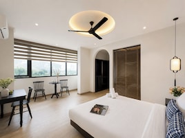 Quarto de hotel moderno com uma cama grande, área de refeições com mesa e cadeiras, ventilador de teto e janelas amplas com persianas.