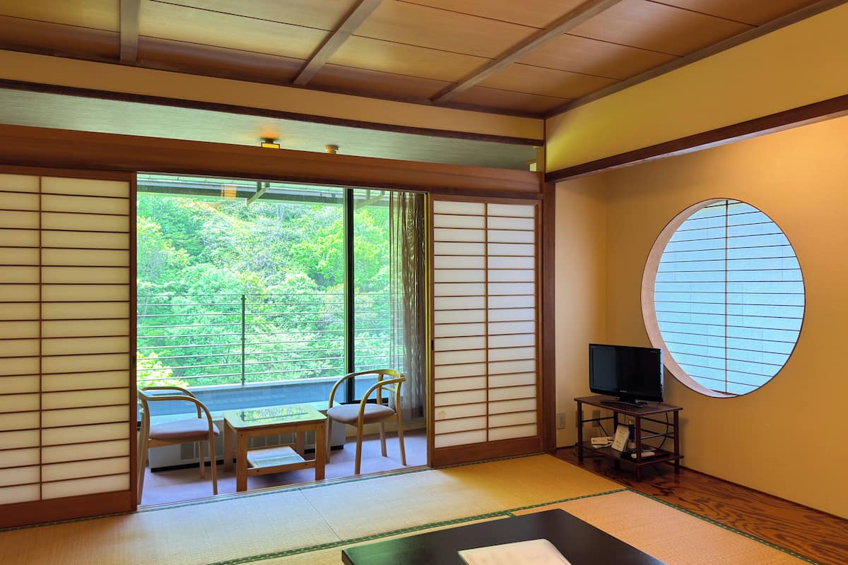 Um quarto japonês tradicional com tatames, uma porta deslizante para uma varanda, uma televisão sobre um suporte e um janelão circular.