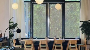 Uma área de refeições com uma mesa comprida, várias cadeiras e janelas amplas com vista para árvores.