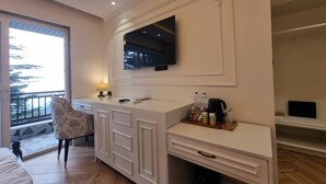 Quarto de hotel com uma janela ampla, televisão de tela plana na parede, escrivaninha com gavetas, uma cadeira e uma mesinha de apoio com produtos de higiene pessoal.