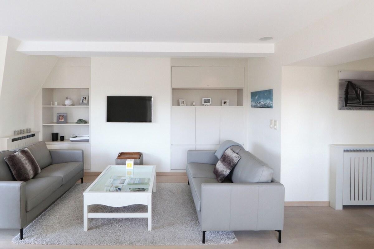 Uma sala de estar moderna com dois sofás cinzentos, uma mesa de centro branca e uma TV fixada na parede.