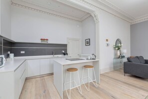 Uma cozinha moderna com armários brancos, uma ilha central com banquetas, um sofá cinzento e um espelho decorativo.