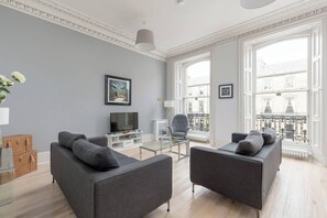 Uma sala de estar moderna com dois sofás cinza, uma mesa de centro de vidro, uma TV de tela plana e uma grande janela com vista para edifícios.
